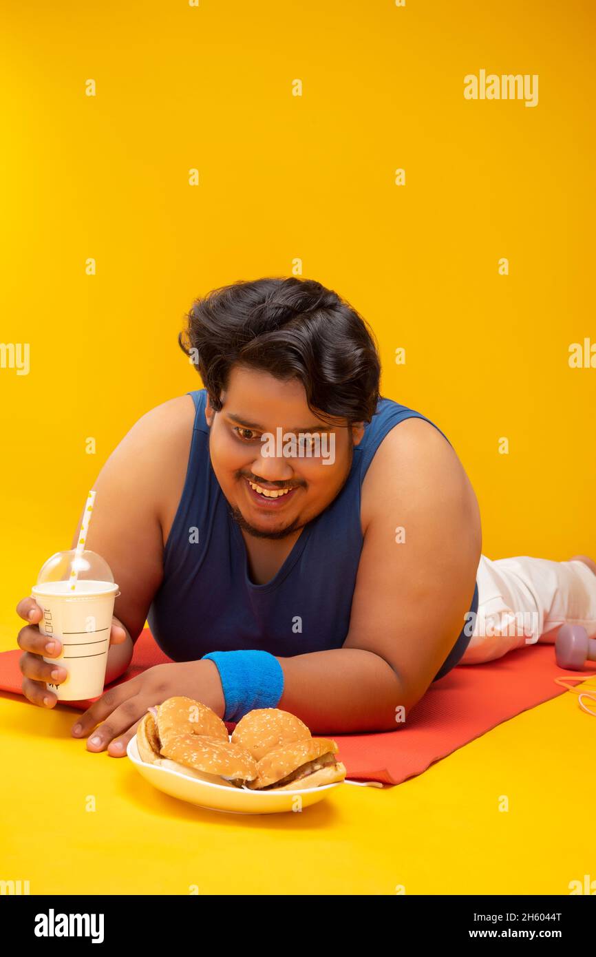 A fat man lying on yoga mat holding a drink greedily devouring at the ...