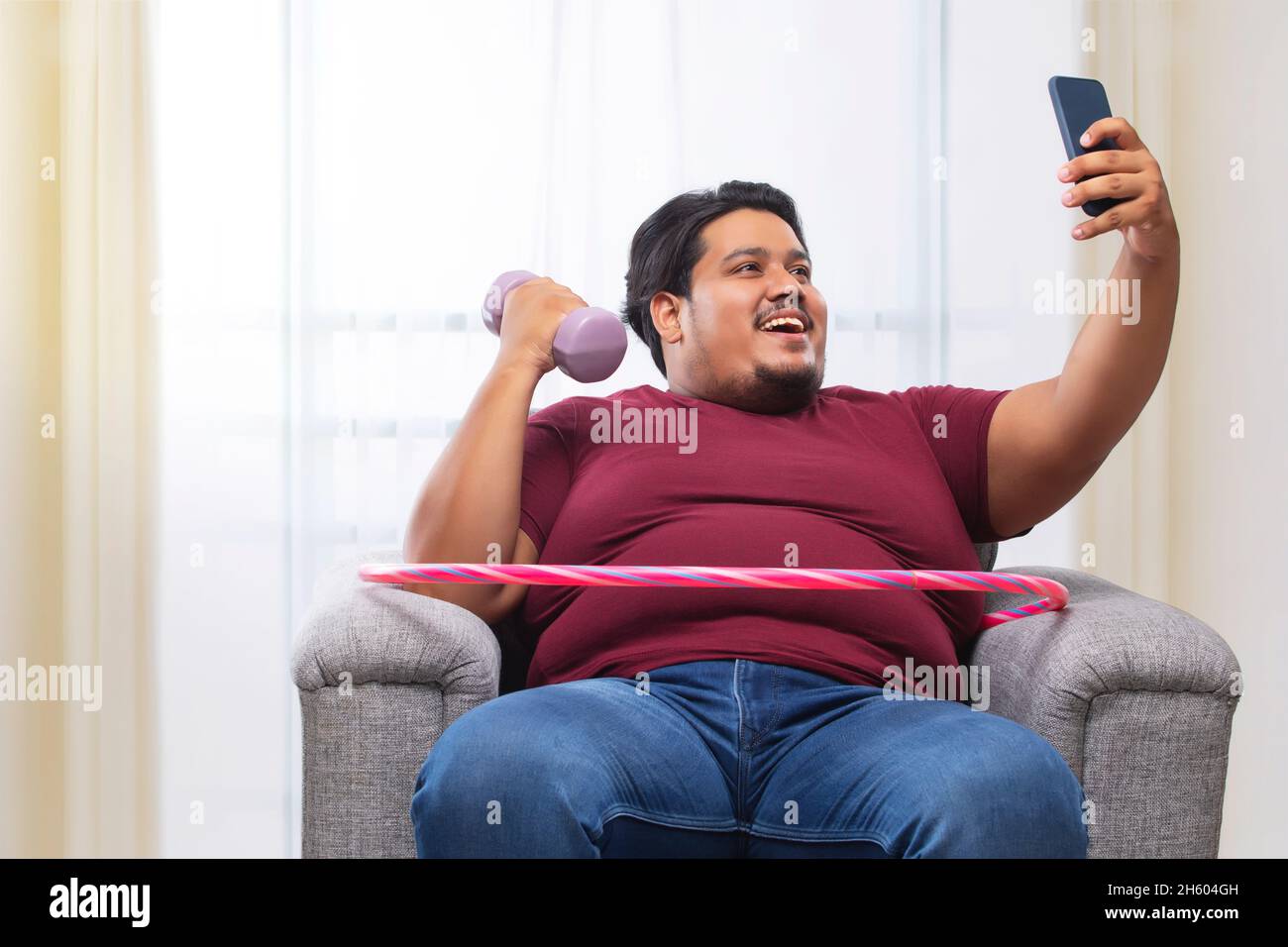 A fat man sitting on couch with dumbles and hulla hoop around his waist ...
