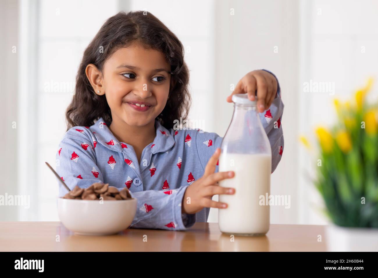 A girl opening cap of a milk bottle to pour into a bowl full of chocos ...