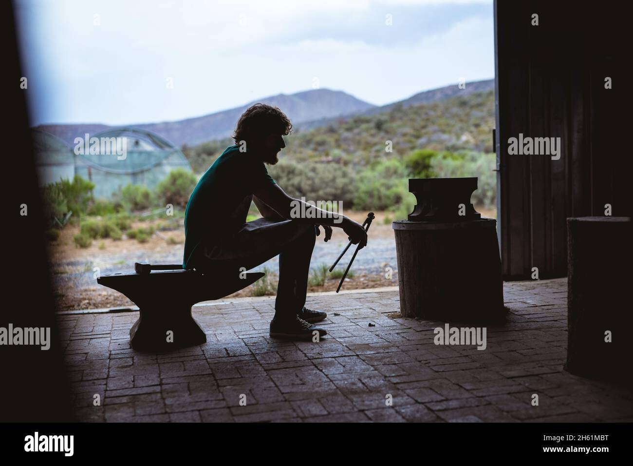 Side view of blacksmith holding work tool while sitting on anvil in ...