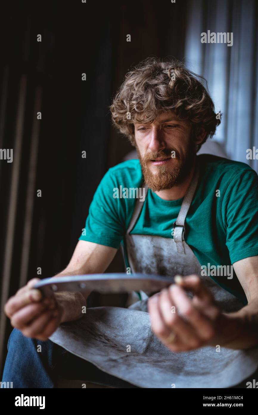 Bearded caucasian blacksmith holding knife shaped metal while sitting ...