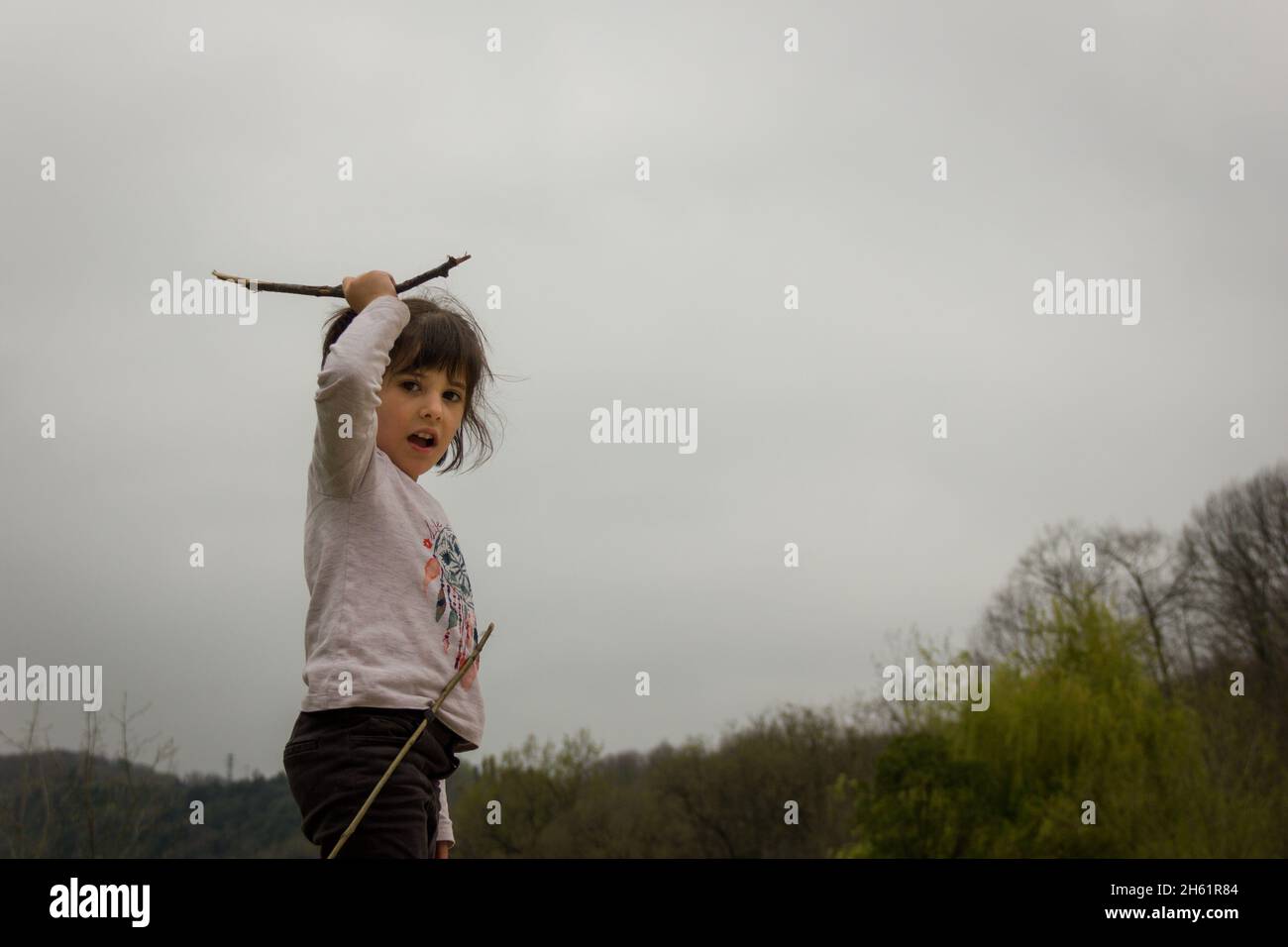 Brave little girl holding a stick with hand up at park on cloudy day in ...