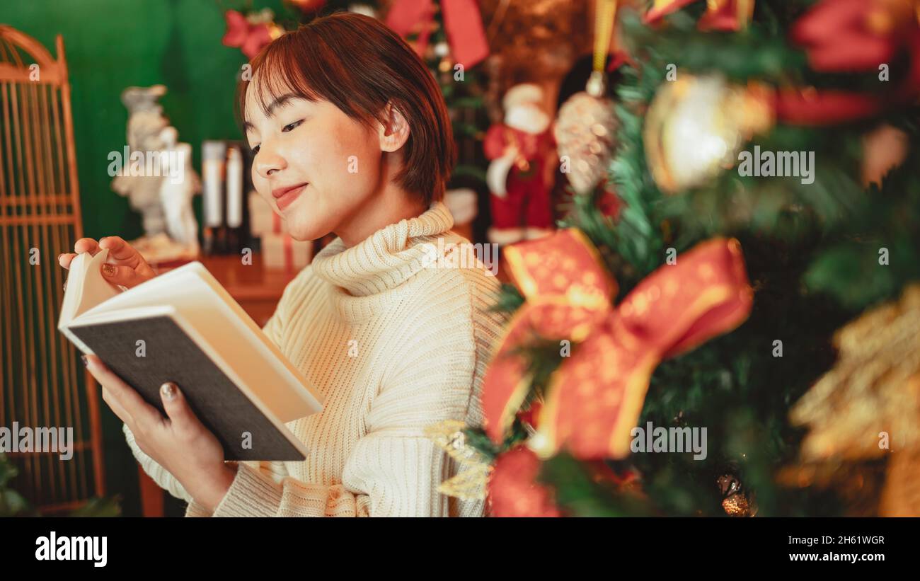a young Woman in front of Christmas tree reading a book at home with x ...