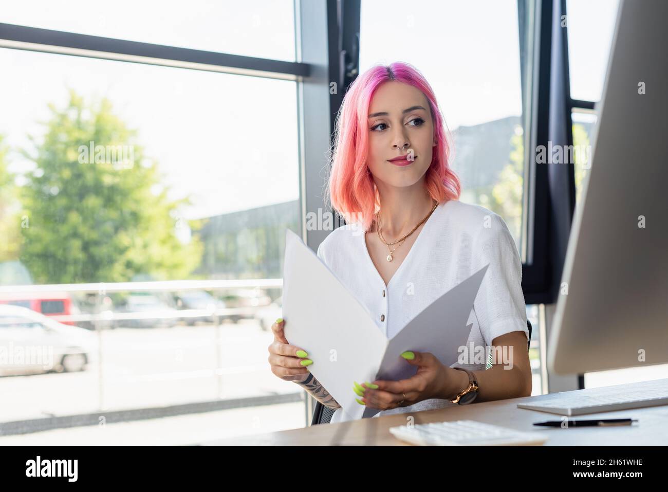 pierced businesswoman with pink hair holding folder and looking at ...
