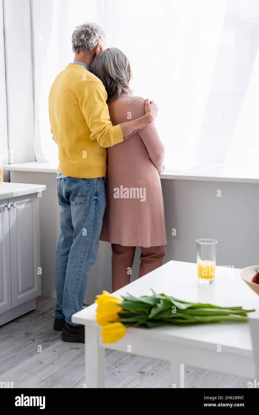 Back view of mature couple hugging near window and blurred tulips on ...