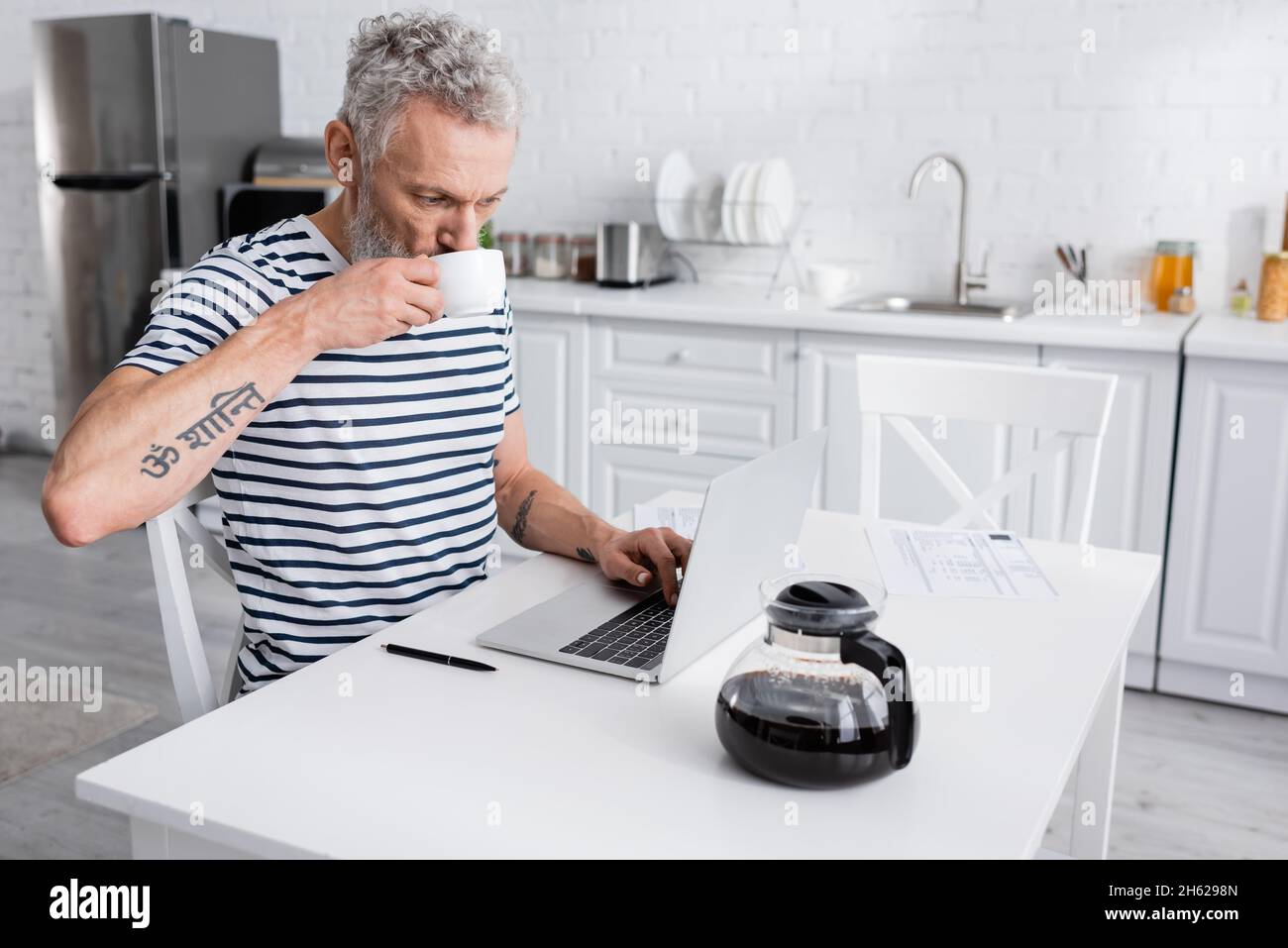 Middle aged man drinking coffee and using laptop near papers in kitchen ...