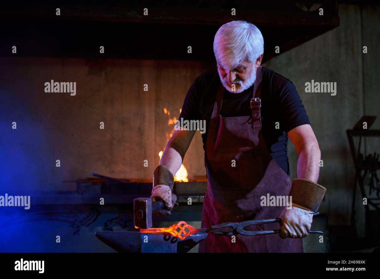 Caucasian man with grey hair and beard processing molten metal at forge ...