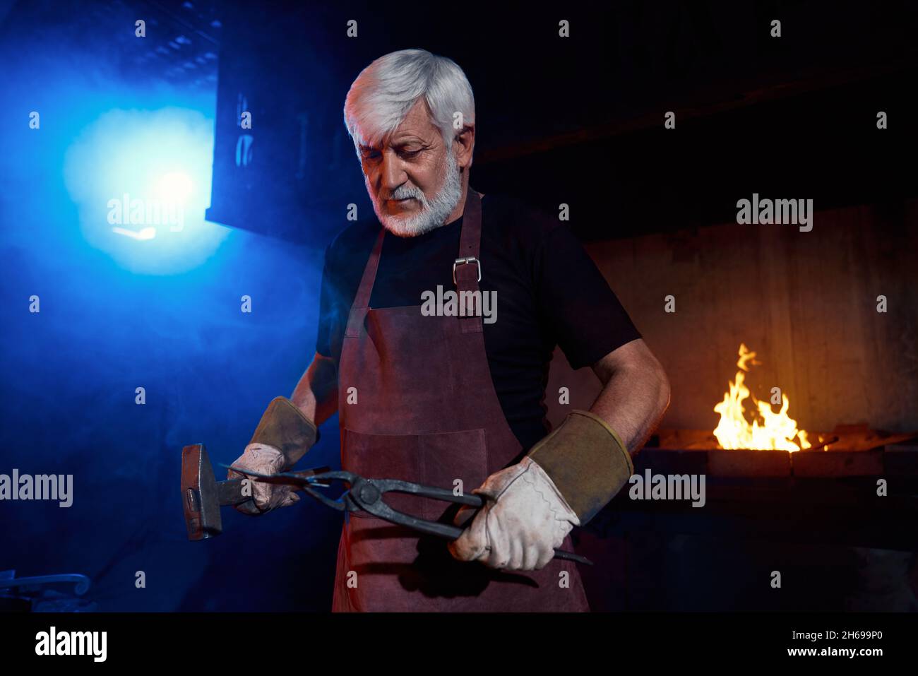 Aged caucasian blacksmith in protective apron and gloves using hammer ...