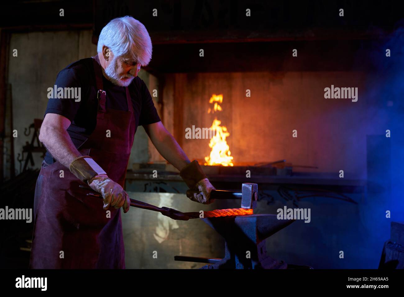 Caucasian grey haired man shaping molten metal on anvil with hammer and ...