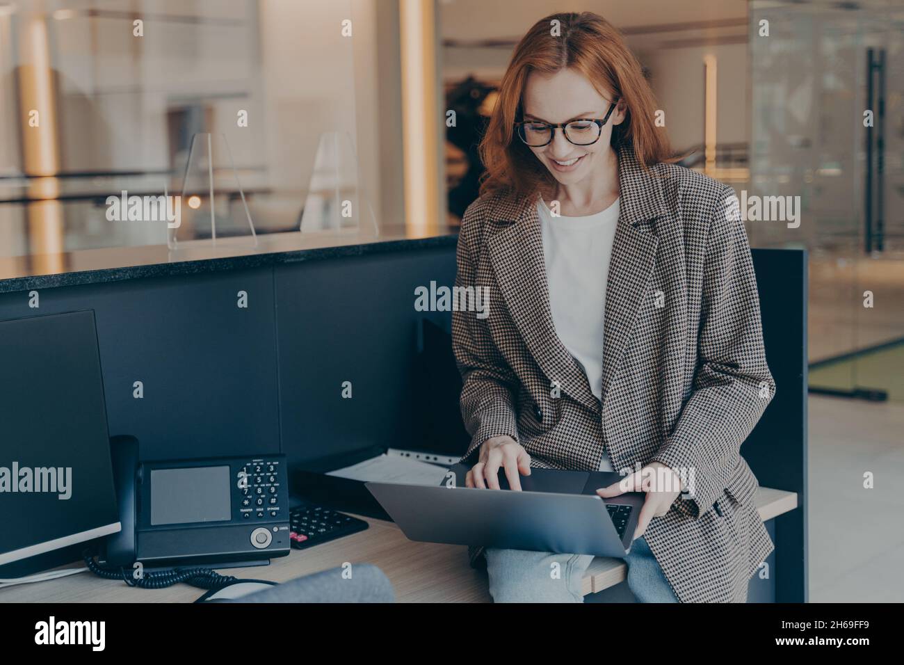 Beautiful redhead businesswoman sitting on top of desk and using laptop ...