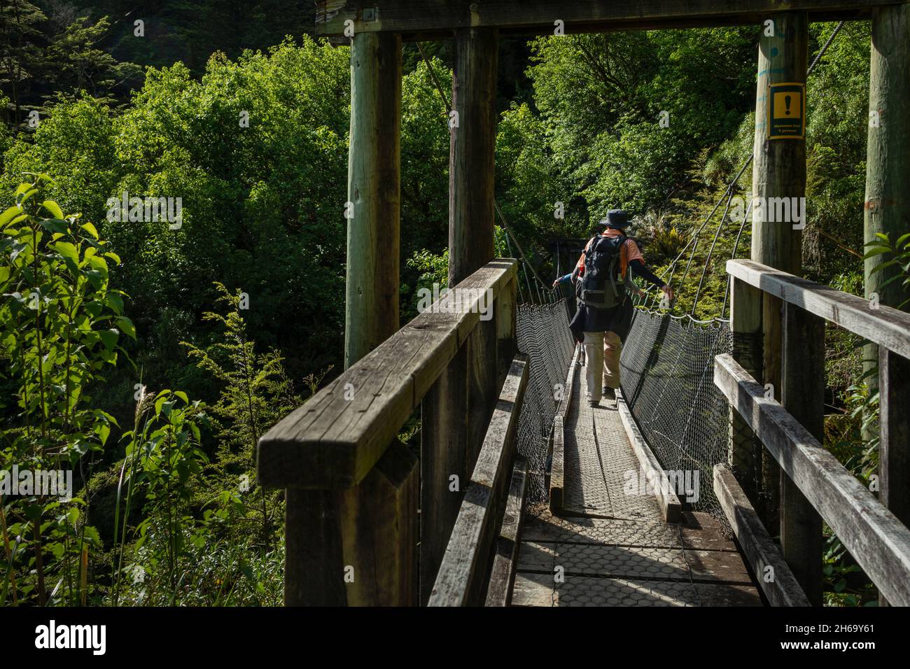 Hikers walking on a swing bridge at Roueturn Track. Yellow warning sign ...