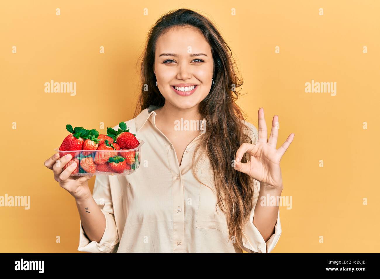 Young hispanic girl holding strawberries smiling positive doing ok sign ...