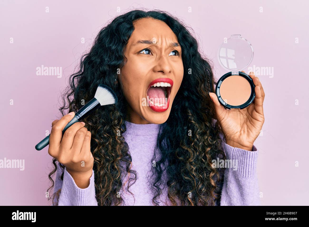 Young latin woman holding makeup brush and blush angry and mad screaming frustrated and furious ...