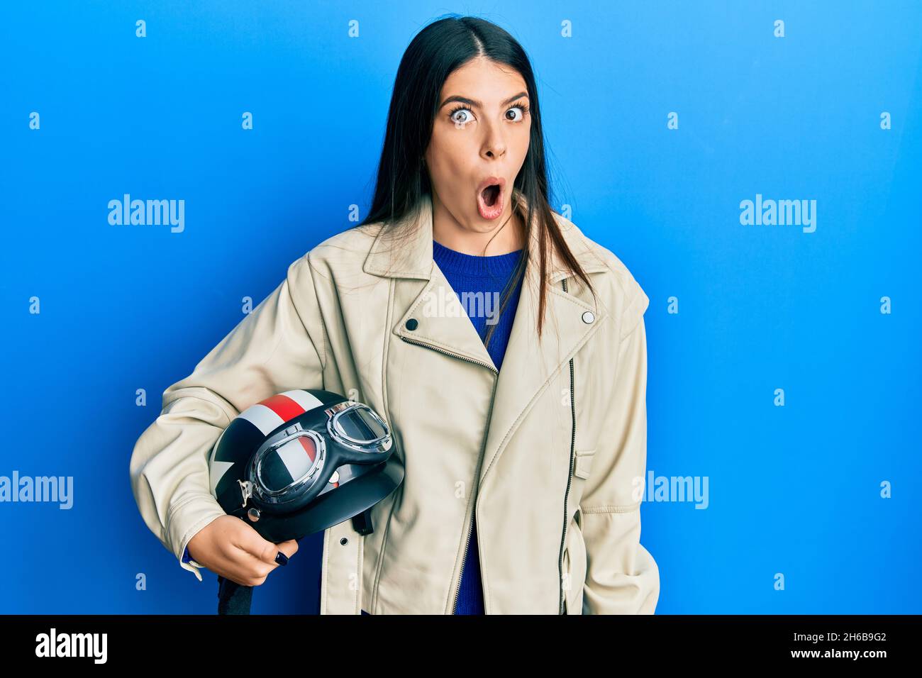 Young hispanic woman holding motorcycle helmet scared and amazed with ...