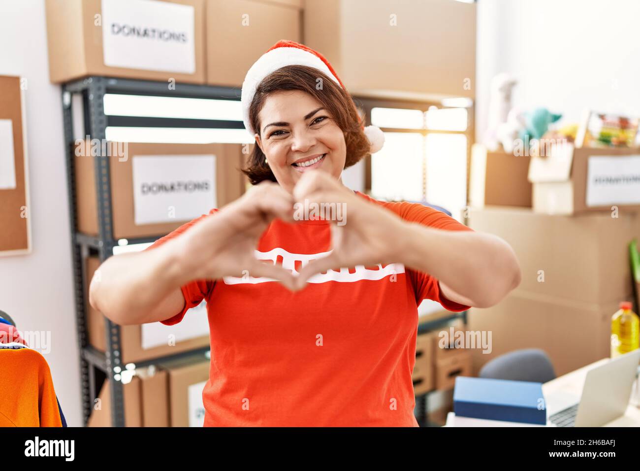 Middle age hispanic woman wearing volunteer t shirt and christmas hat smiling in love showing ...