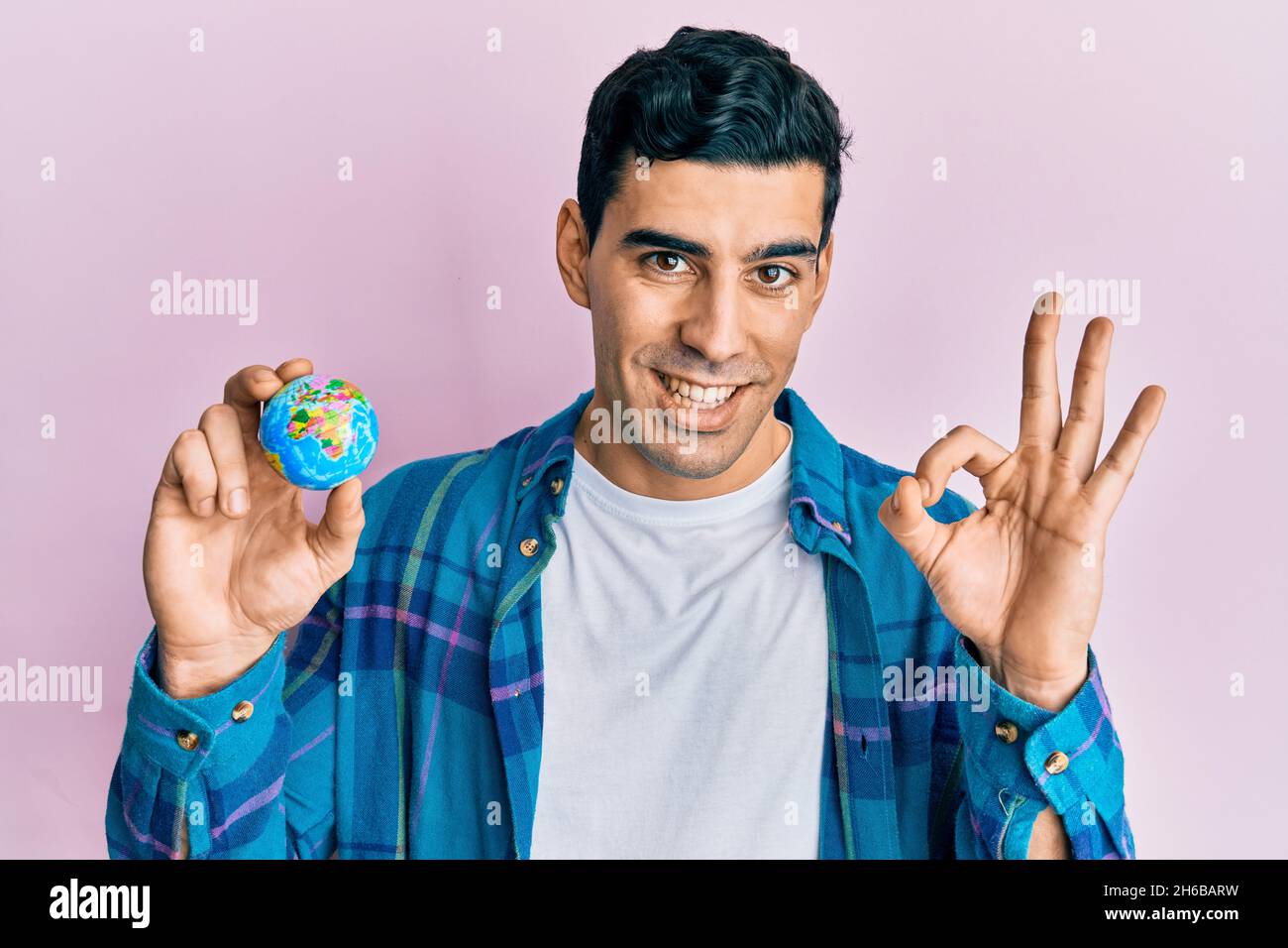 Handsome hispanic man holding small world ball doing ok sign with ...