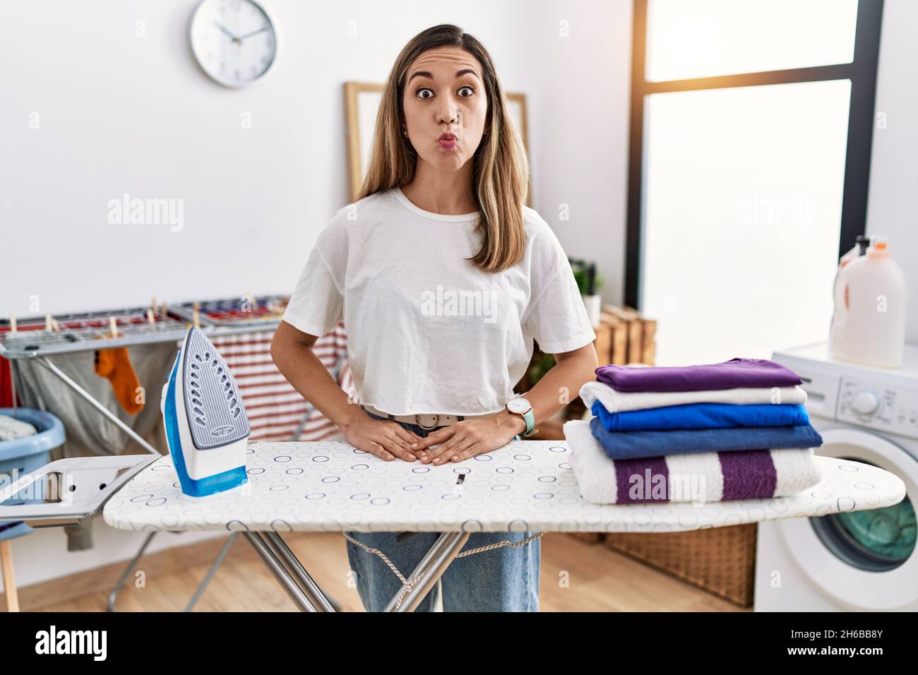 Young hispanic woman ironing clothes at laundry room puffing cheeks ...