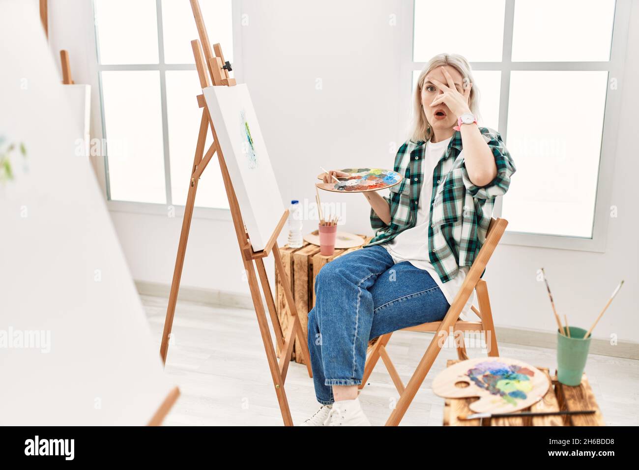 Young artist woman painting on canvas at art studio peeking in shock ...