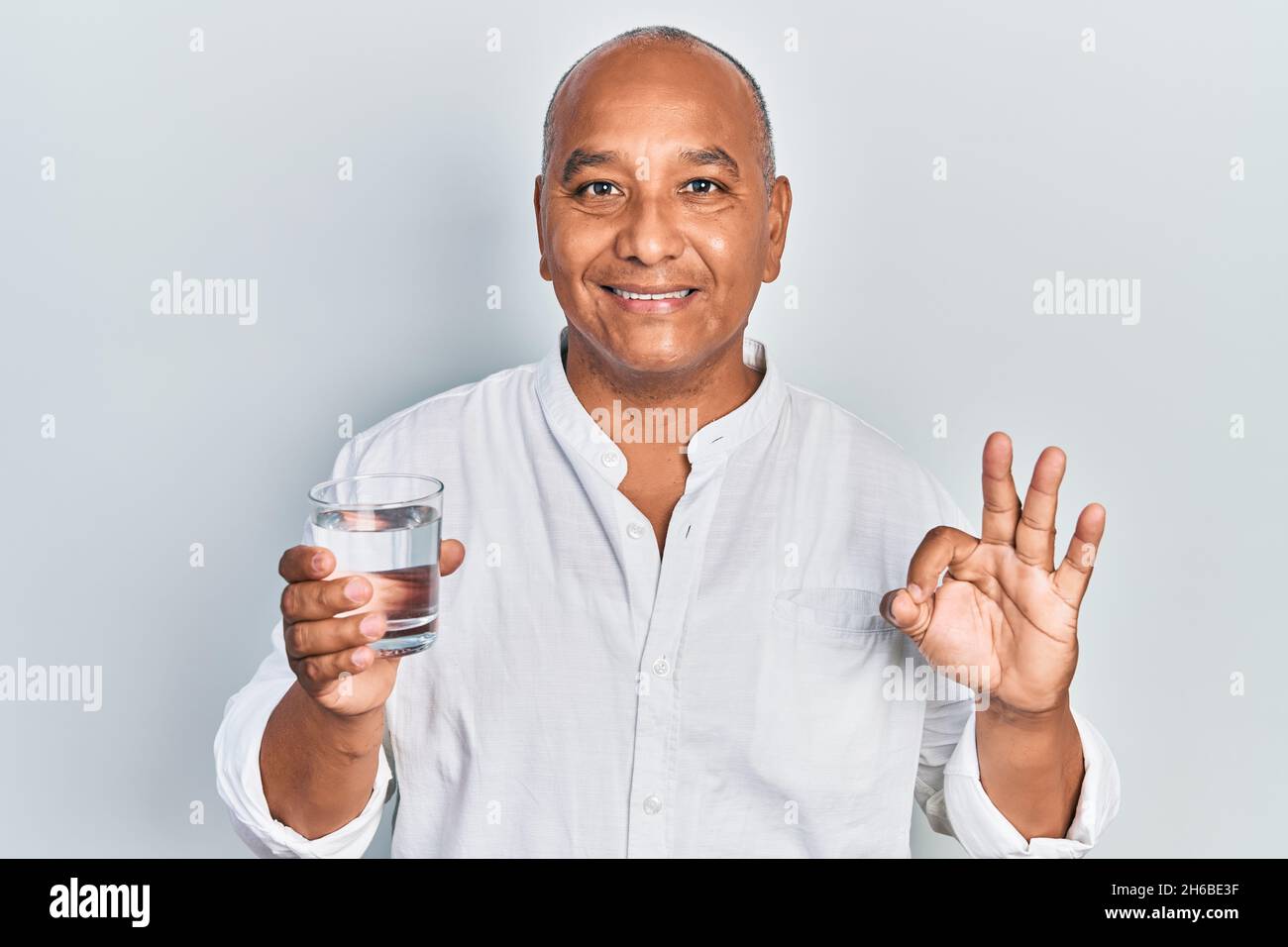 Middle age latin man drinking glass of water doing ok sign with fingers ...