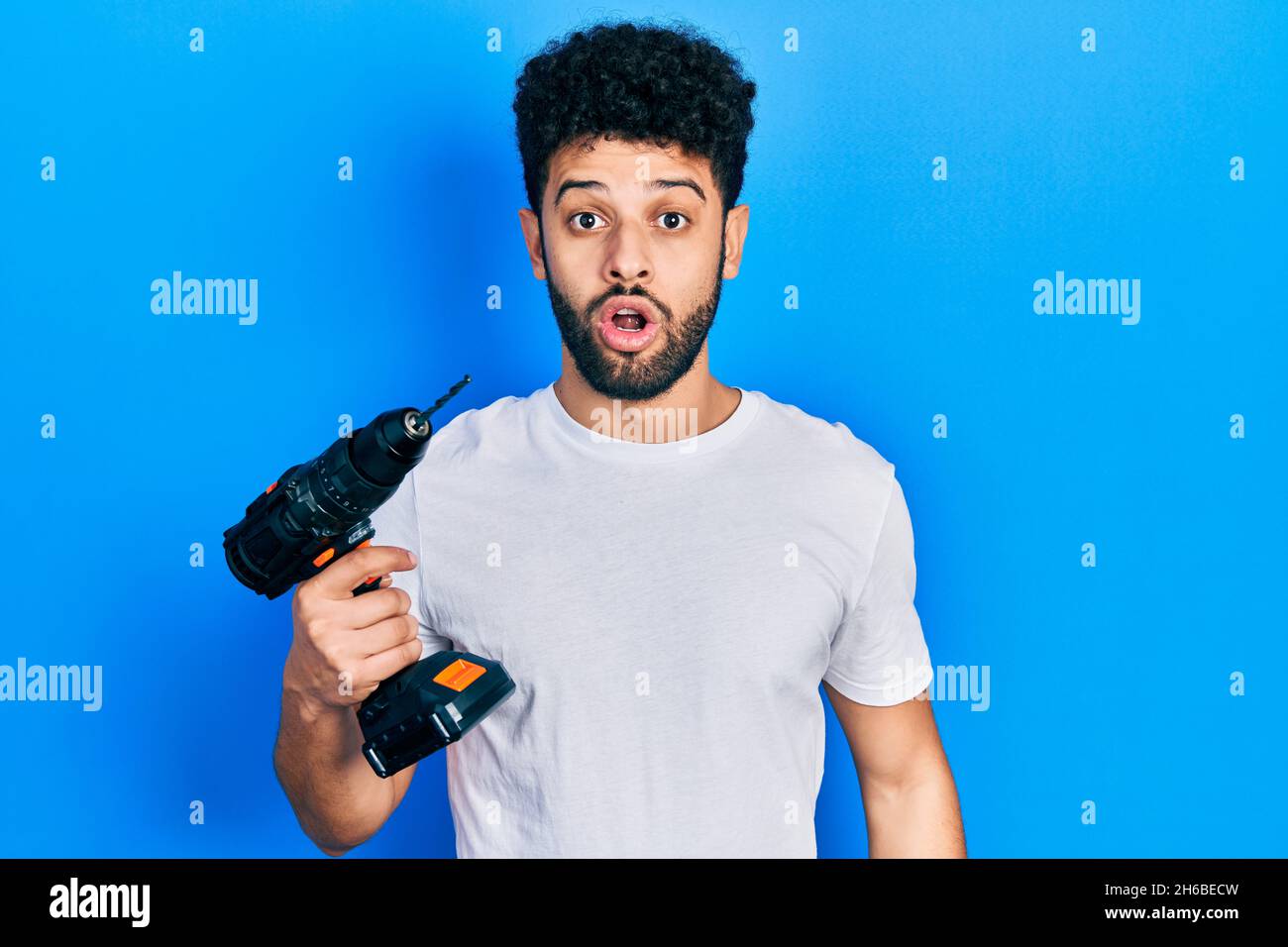 Young arab man with beard using drill scared and amazed with open mouth ...