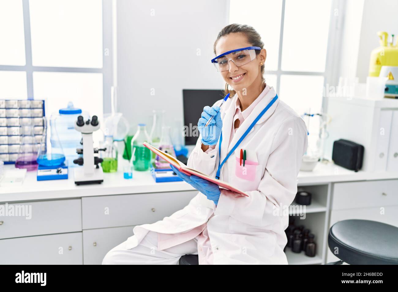 Young hispanic woman wearing scientist uniform writing on notebook at ...