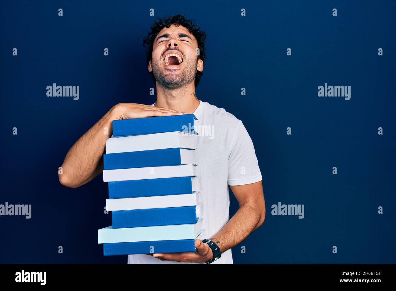 Young hispanic man holding a pile of books angry and mad screaming ...