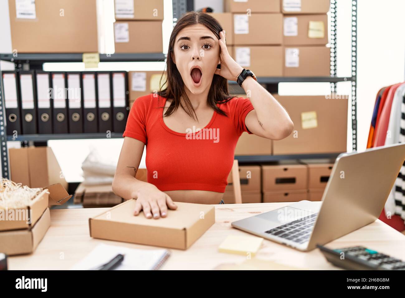 Young hispanic woman preparing order working at storehouse crazy and scared with hands on head ...