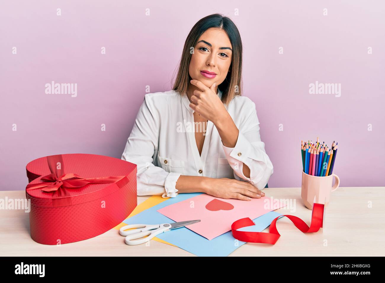 Young hispanic woman making valentine gift sitting on the table looking ...