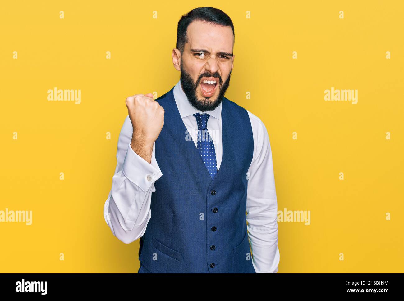 Young man with beard wearing business vest angry and mad raising fist ...