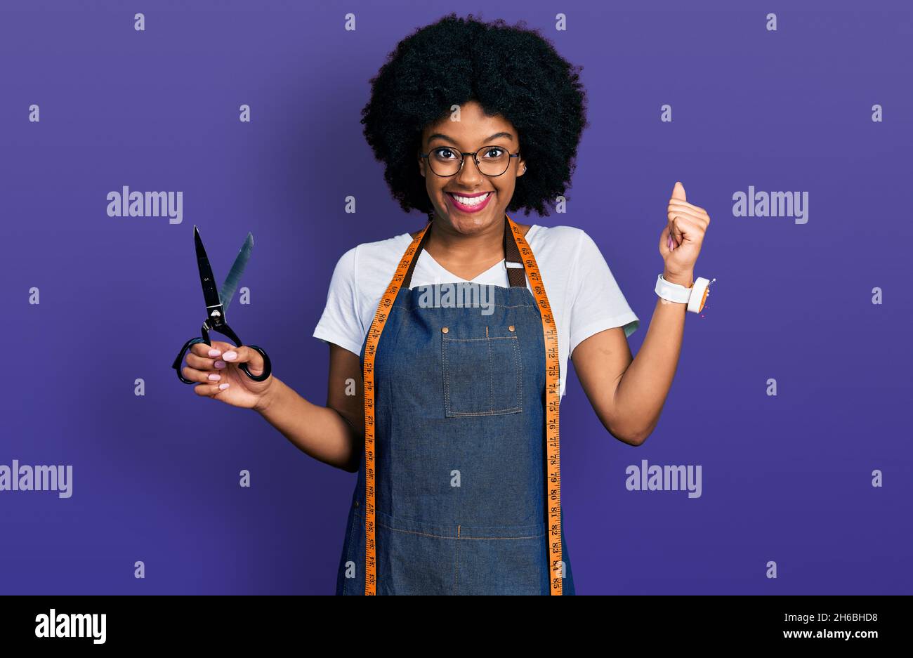 Young african american woman dressmaker designer wearing atelier apron holding scissors ...