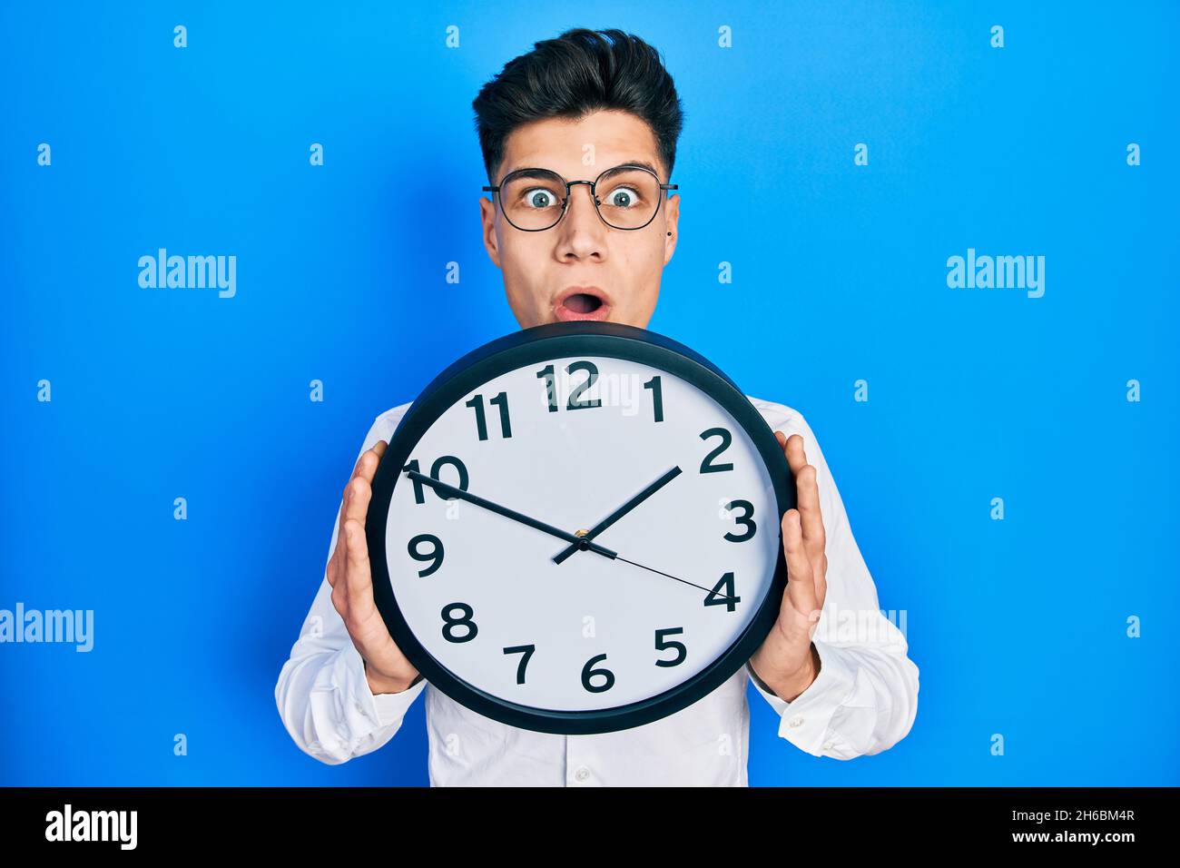 Young hispanic man holding big clock over face afraid and shocked with ...