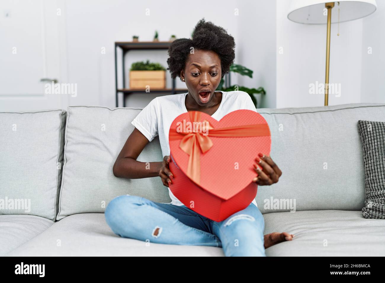 African young woman opening romantic gift afraid and shocked with ...
