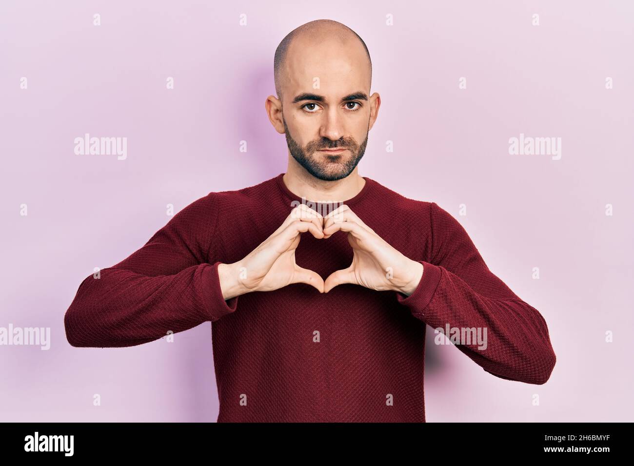 Young bald man doing heart symbol with hands relaxed with serious ...
