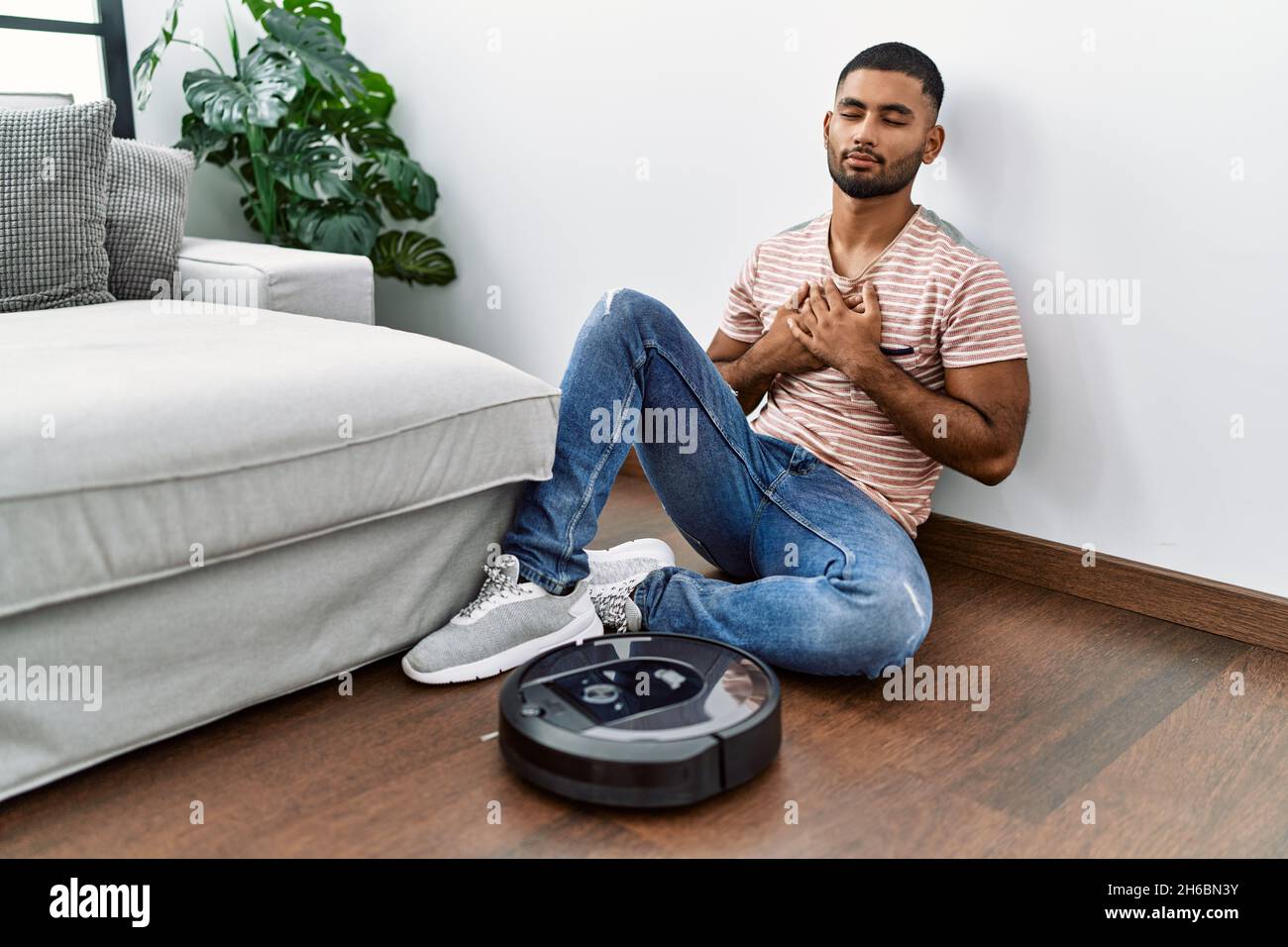 Young indian man sitting at home by vacuum robot smiling with hands on ...