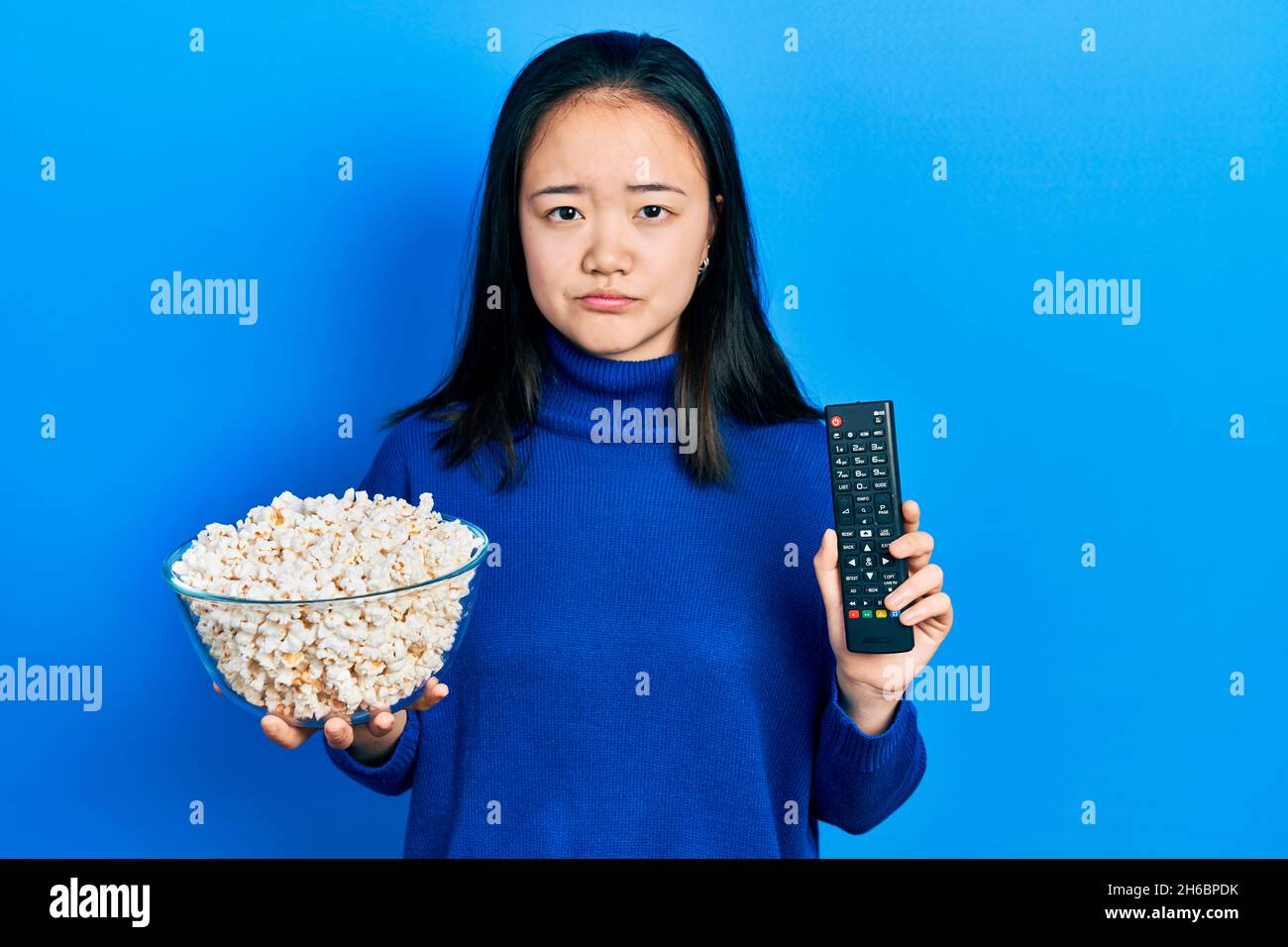 Young chinese girl eating popcorn using tv control depressed and worry ...