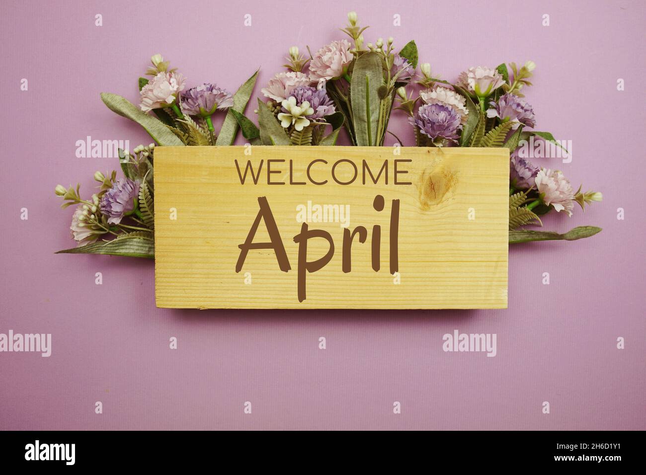 Welcome April text on wooden board with flowers frame on purple ...