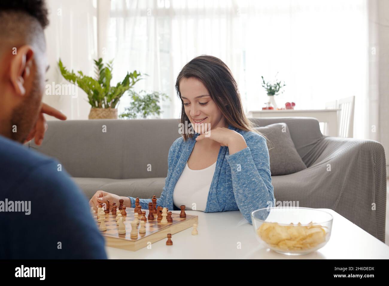 Pretty young woman enjoying playing chess and eating potato chips with ...