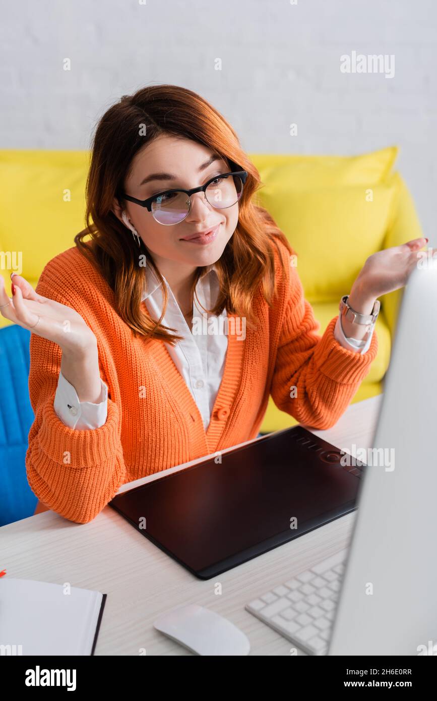 positive woman in earphone showing shrug gesture during video call near ...