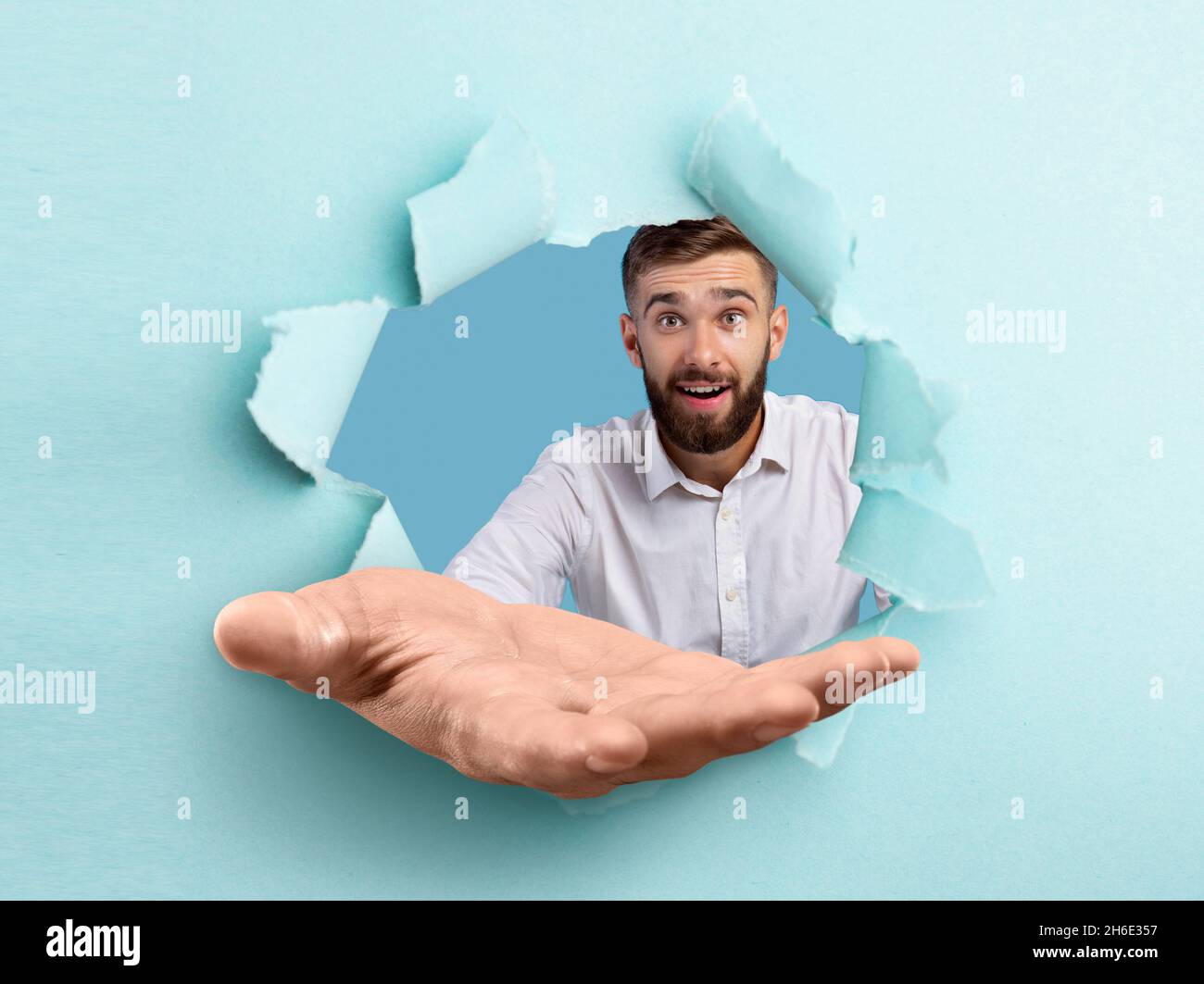 Happy young guy stretching open palm through hole in torn blue paper ...