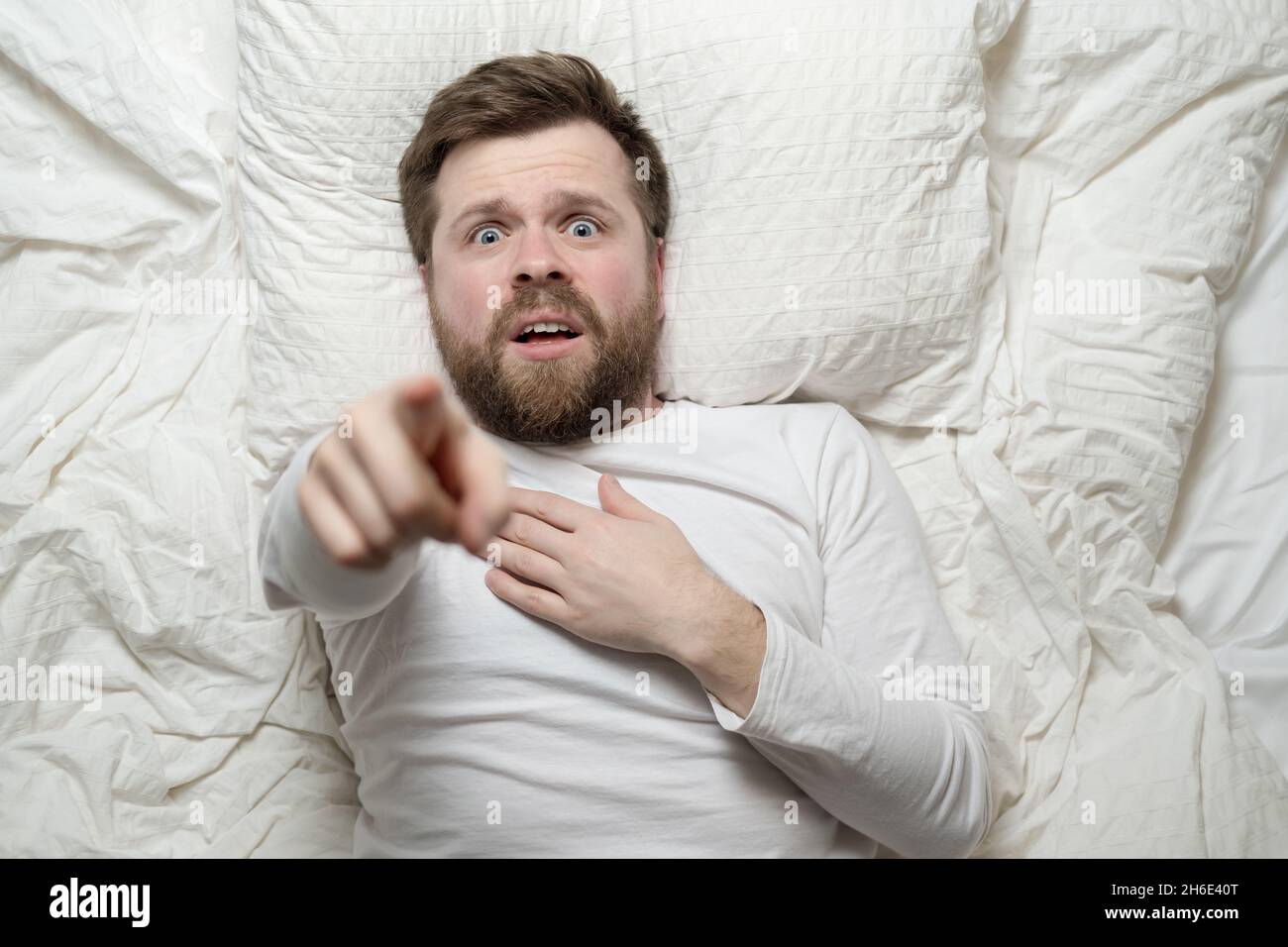 Frightened man lies in bed pointing up and holds hand on chest, he is ...