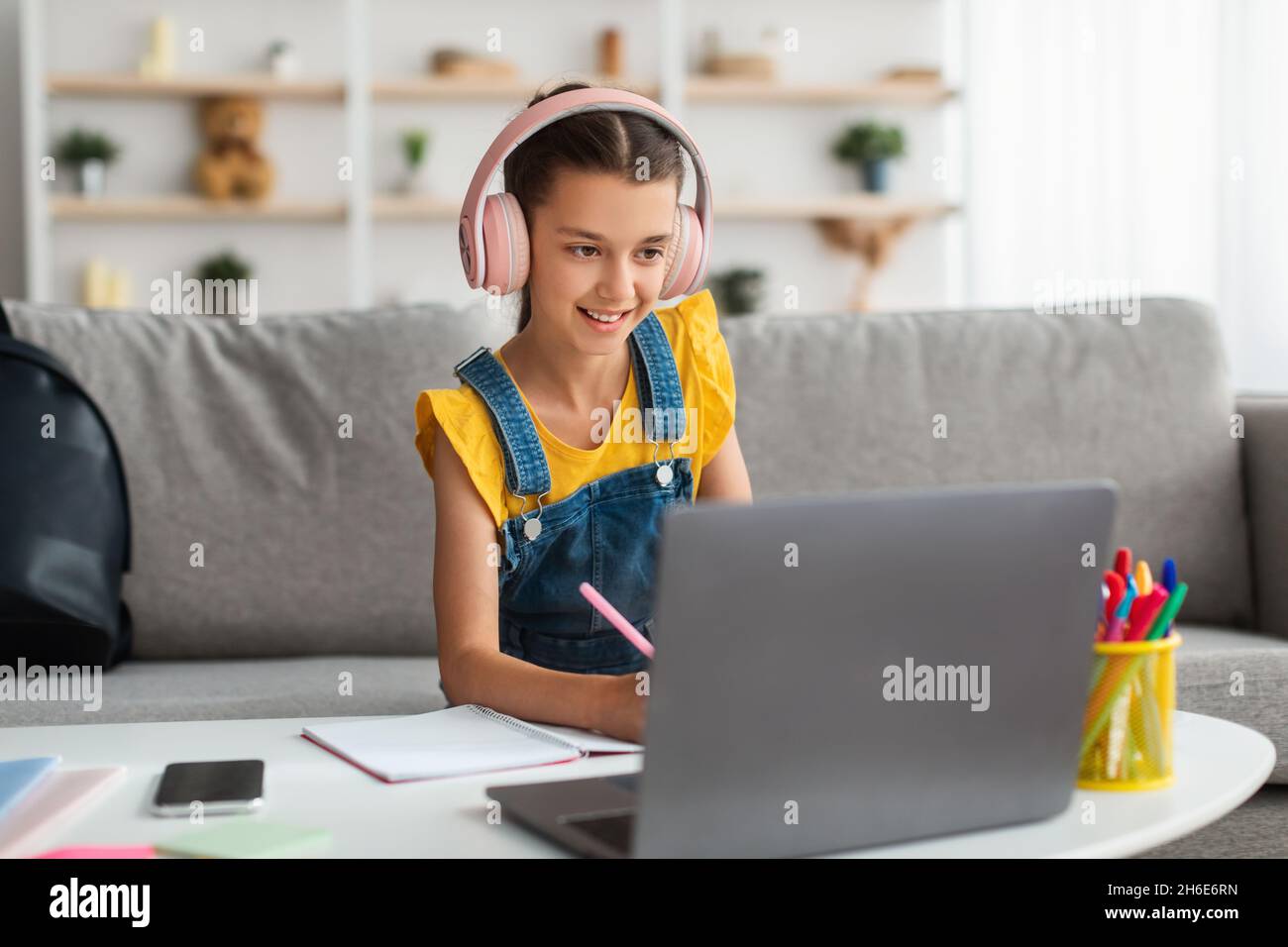 Schoolgirl in wireless headset sitting at desk, writing in textbook ...