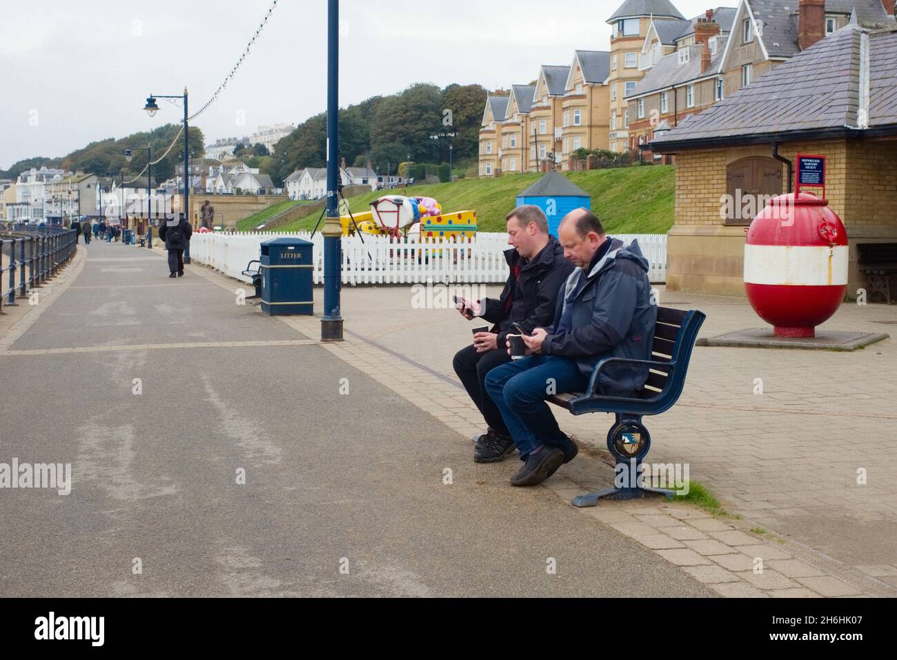 Two men sat on a seaside bench drinking coffees and both looking at ...