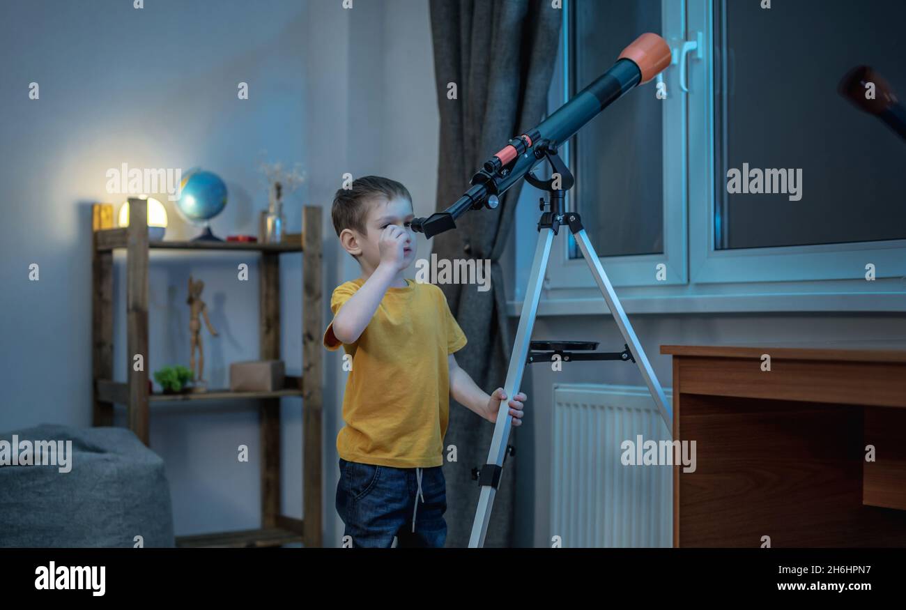Cute boy is looking through a telescope in a room at the night starry ...
