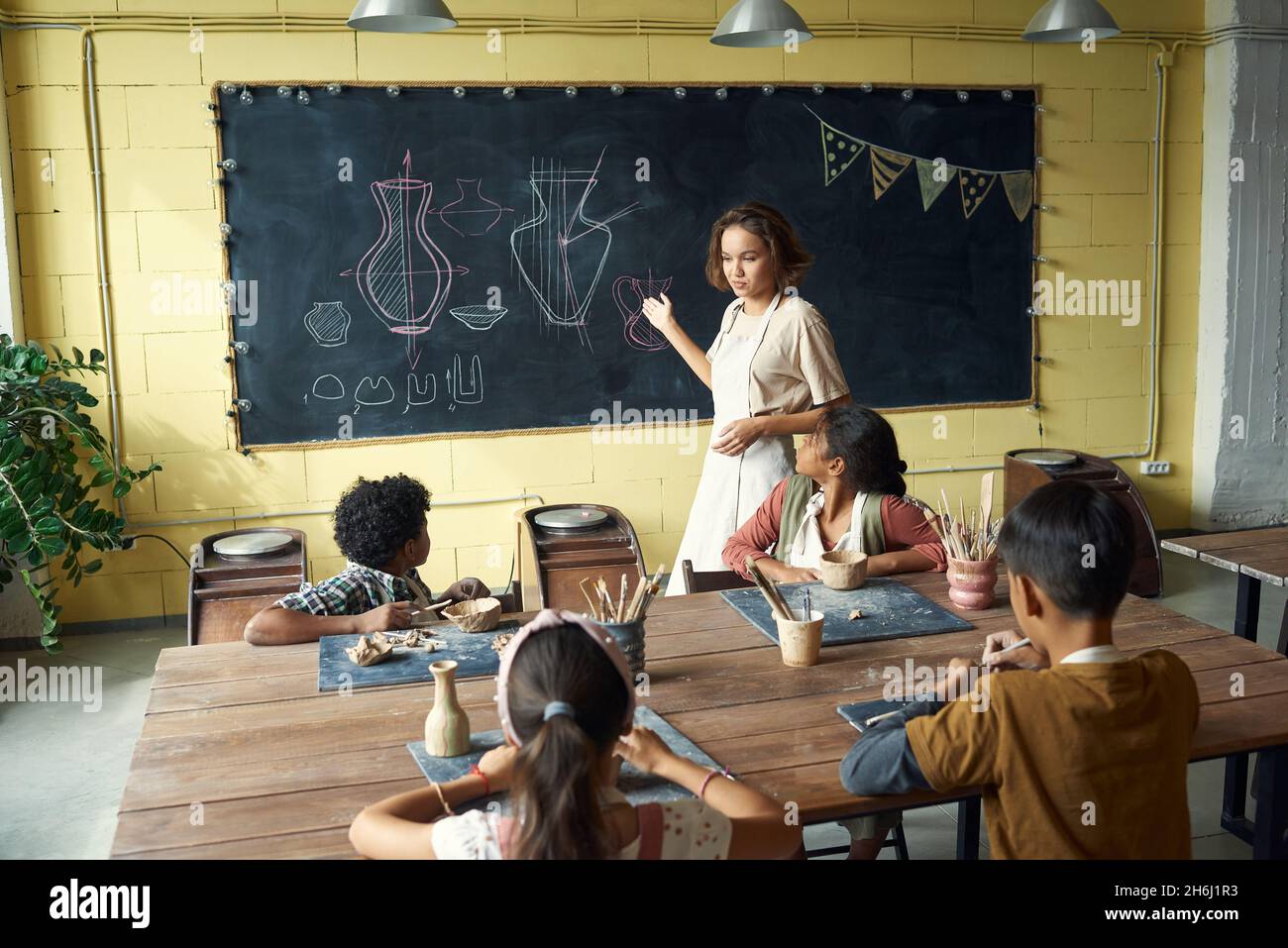 Young confident teacher explaining sketch of pottery to pupils sitting ...