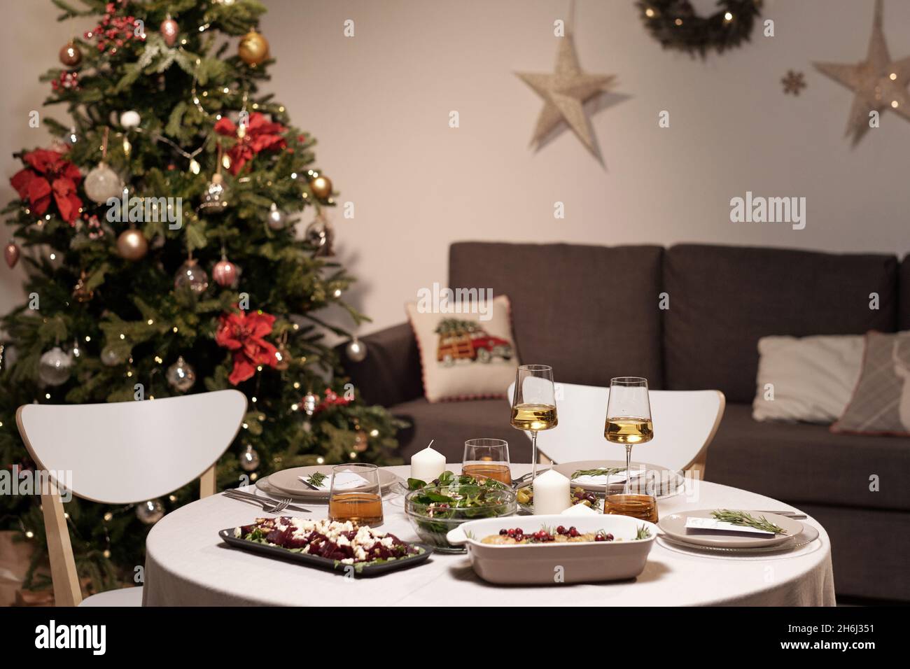 Round table served for festive family dinner on background of decorated xmas tree and couch with ...
