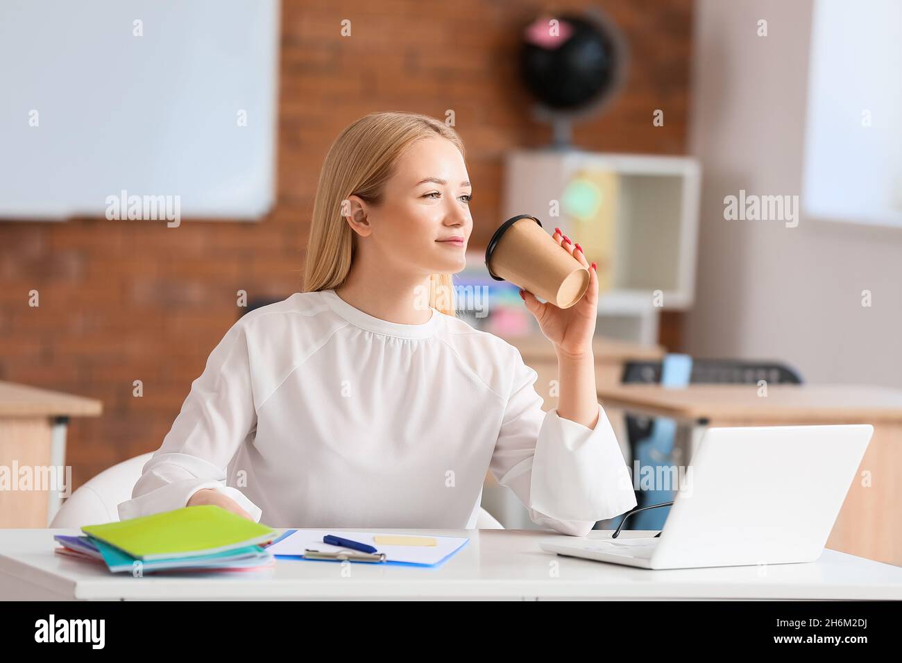 Young female teacher drinking coffee at desk in classroom Stock Photo ...