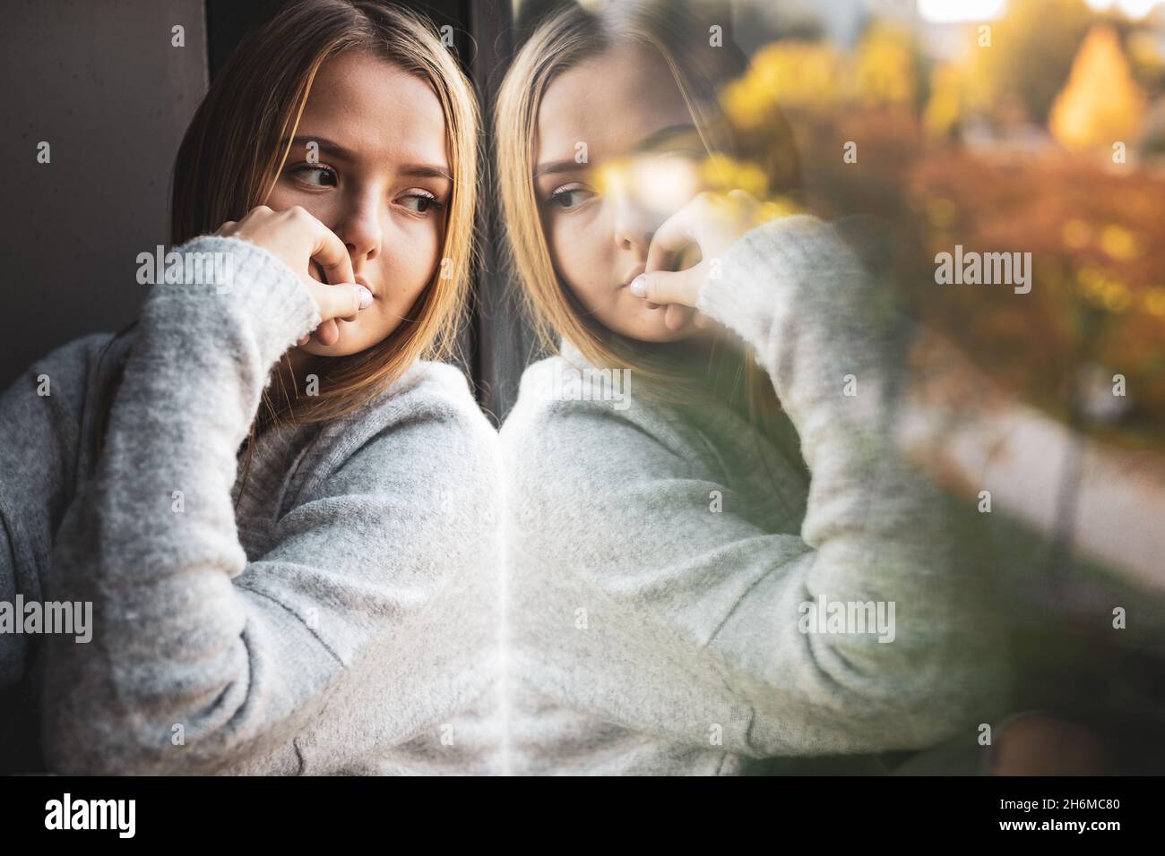 Depressed and anxious young woman sitting by a large window, feeling ...