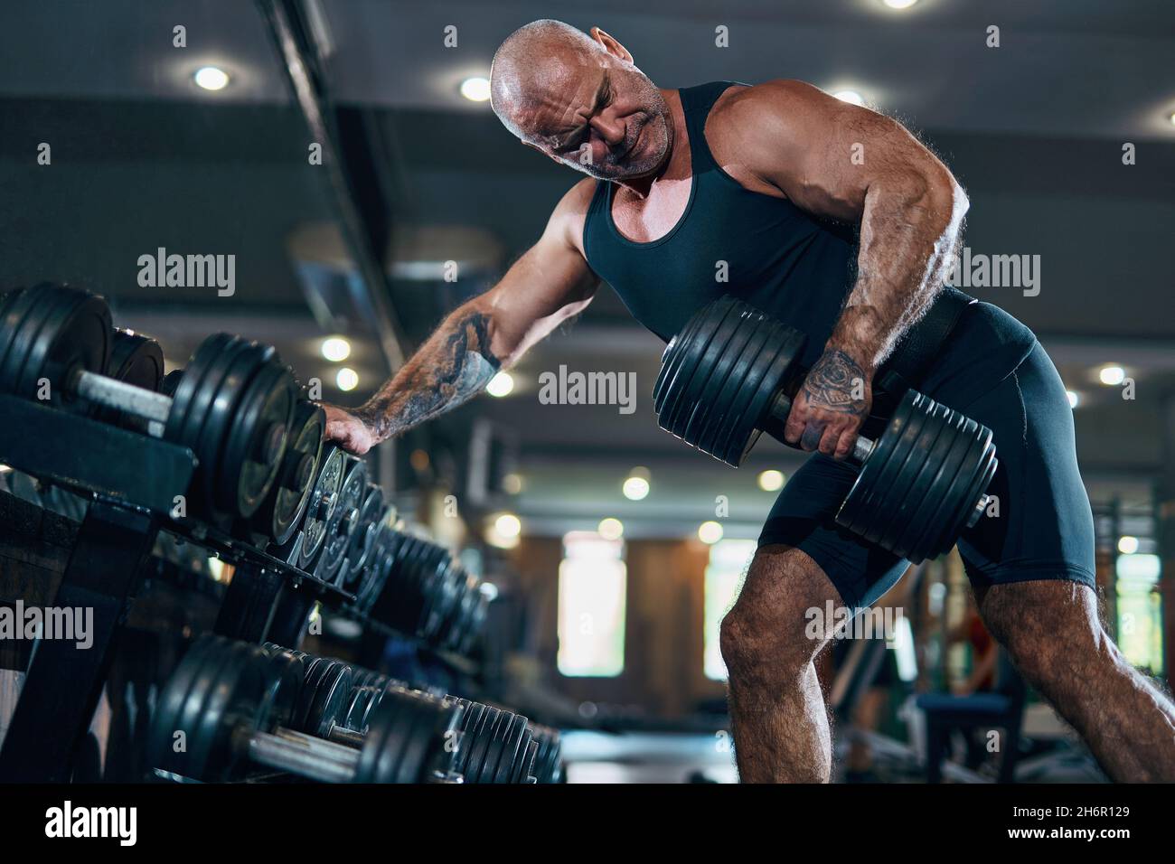 Aged man bodybuilder doing exercises with dumbbells while posing in gym ...