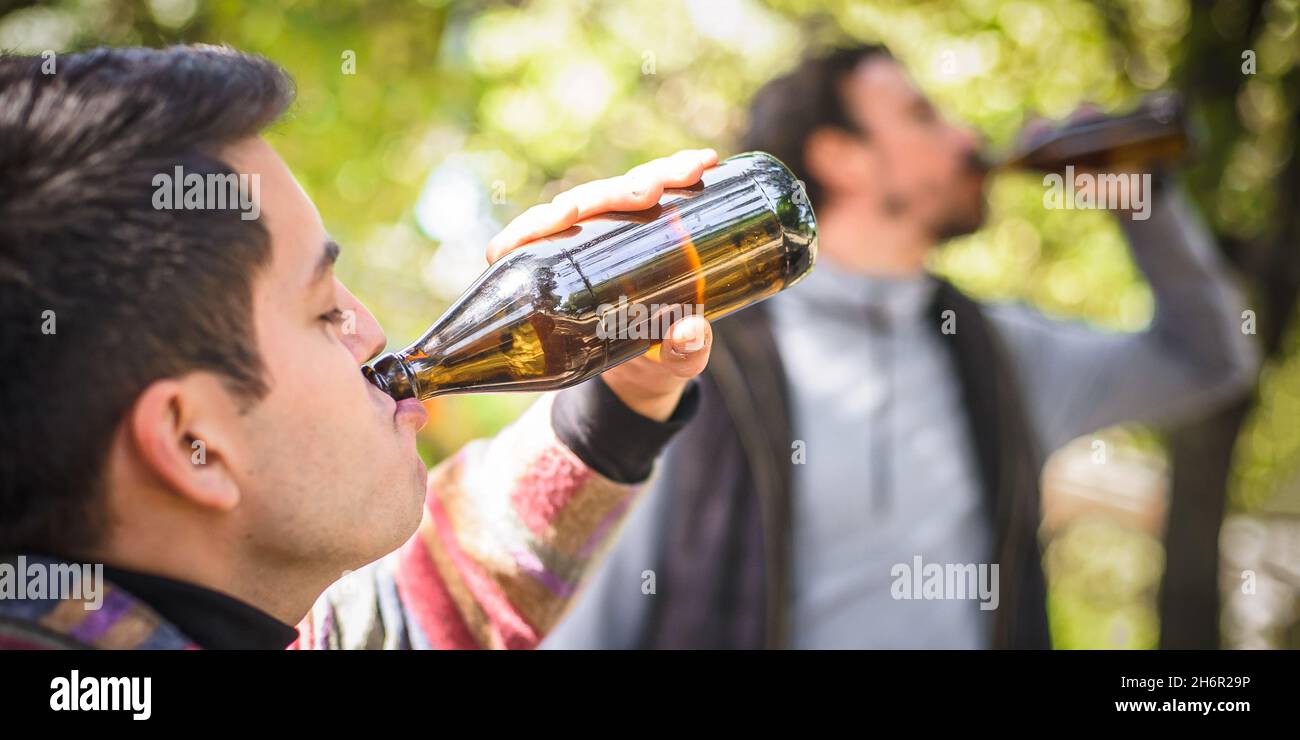 Two men chugging beer from bottle on wooden bench with table in forest ...