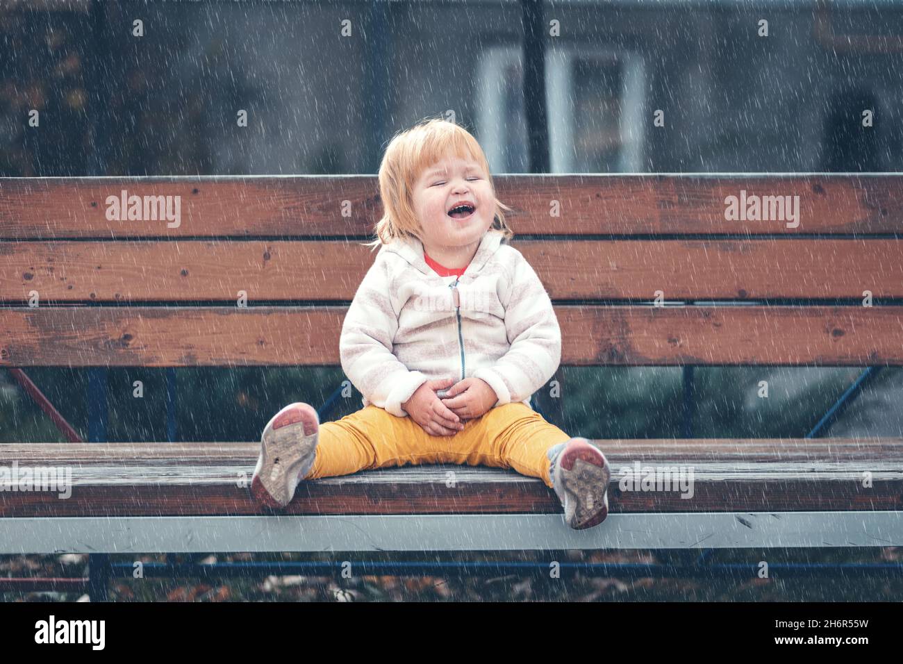 A sad little child is sitting on a bench on the playground alone. Rainy ...